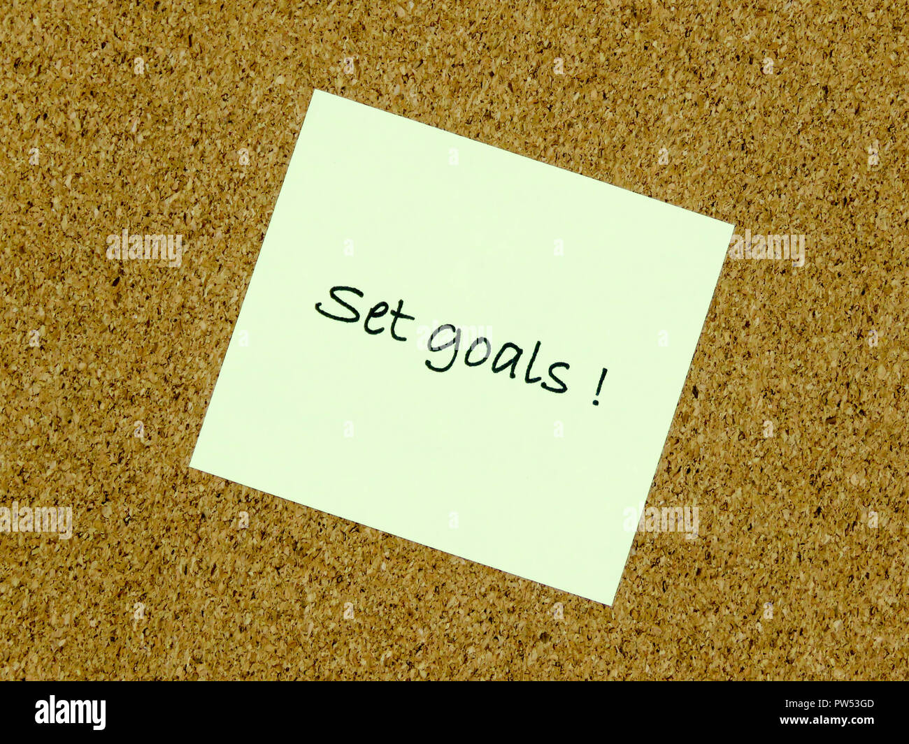 A yellow sticky note with set goals written on it on a cork board ...