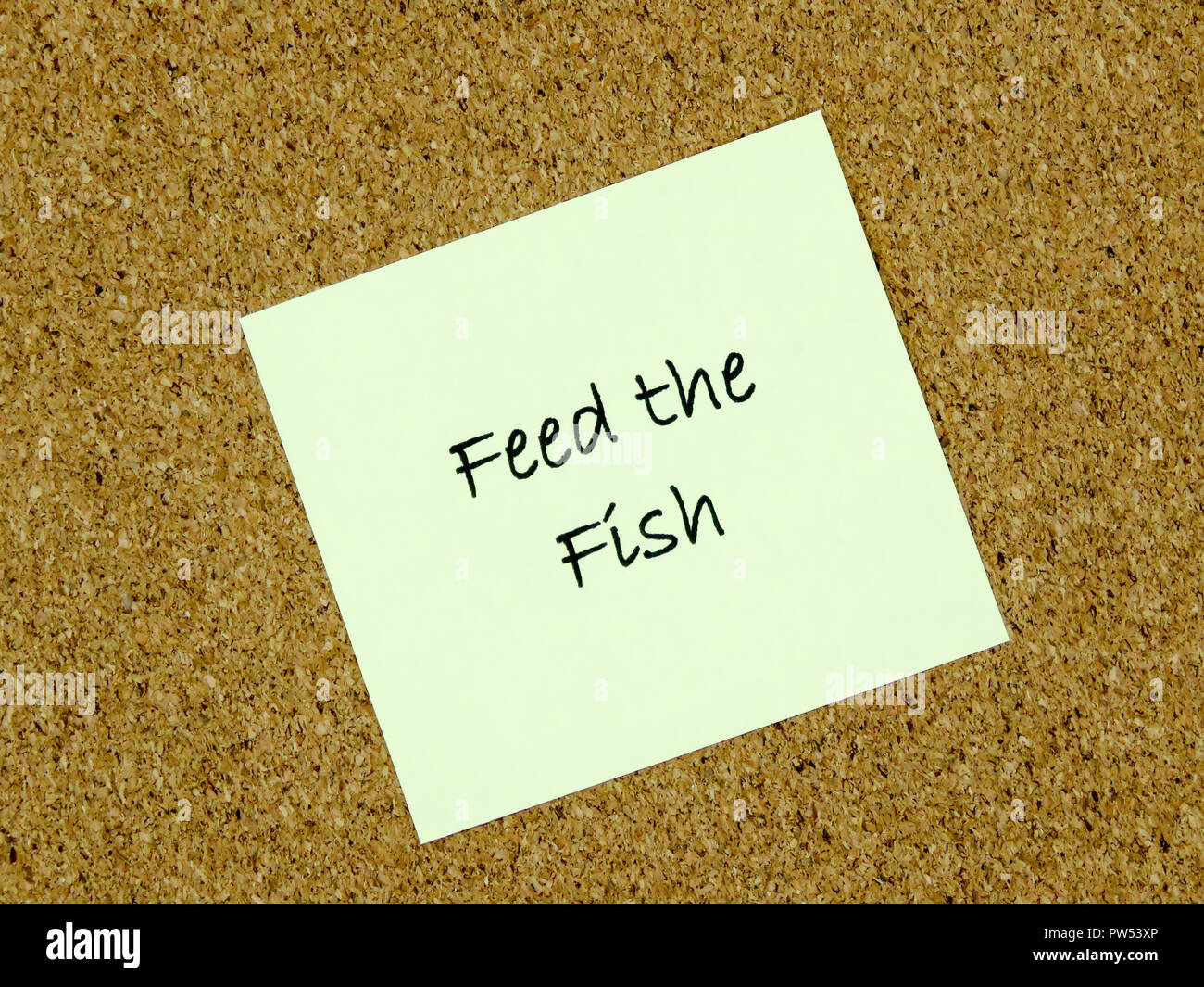 A yellow sticky note with feed the fish written on it on a cork board ...