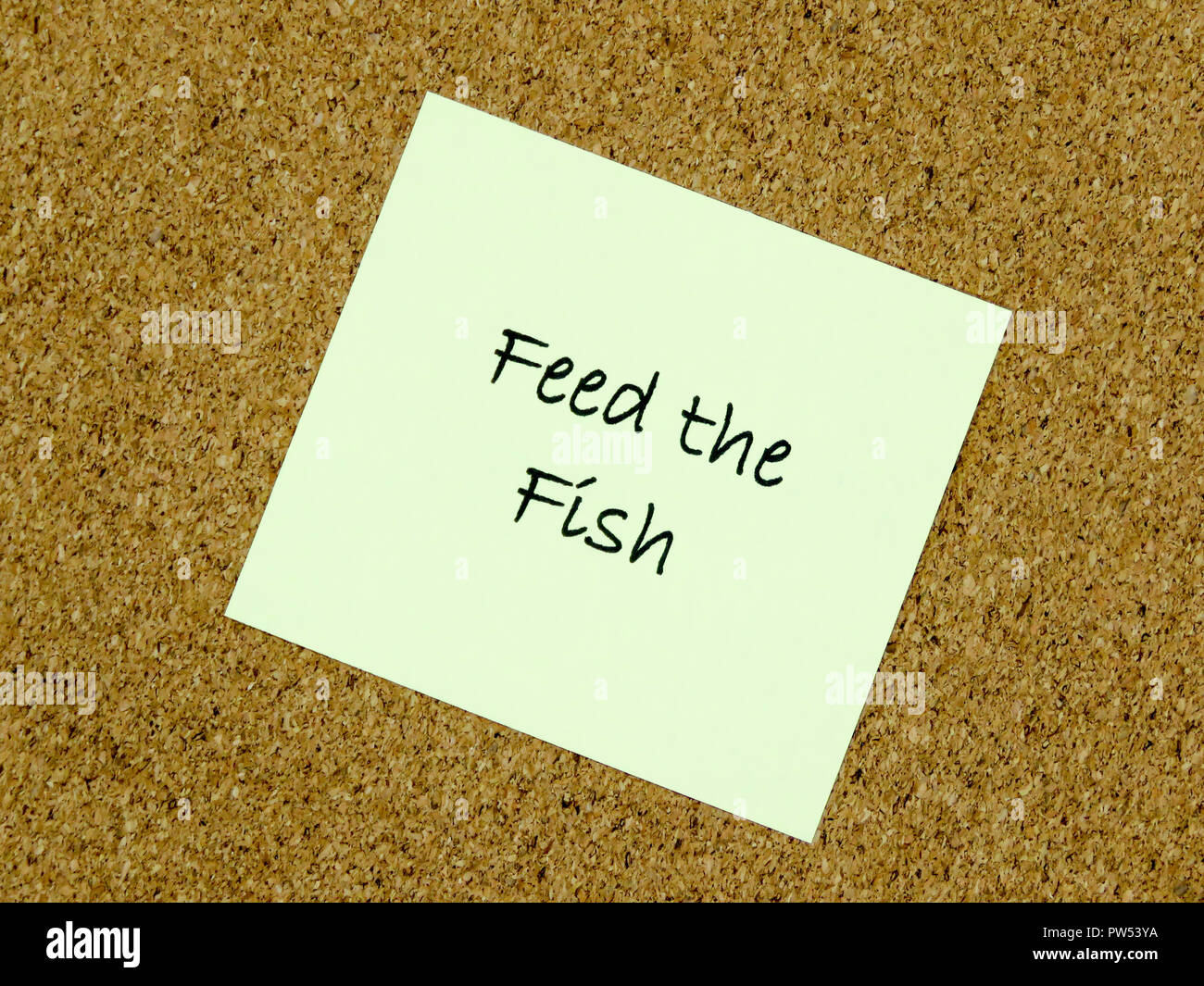 A yellow sticky note with feed the fish written on it on a cork board ...