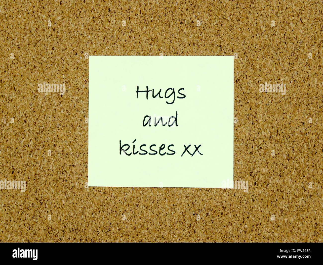 A yellow sticky note with hugs and kisses written on it on a cork board ...