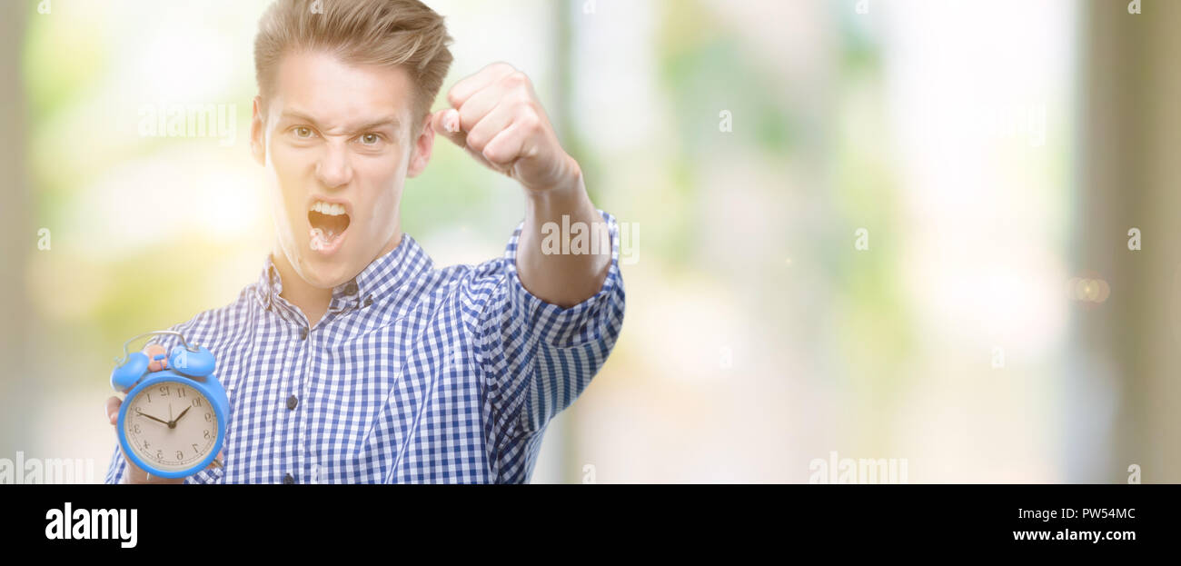 Young handsome blond man holding alarm clock annoyed and frustrated ...