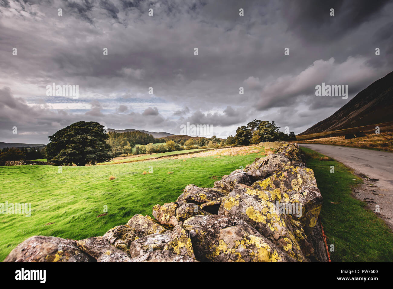 Stone wall aside rural road leading to group of trees, mountains and ...
