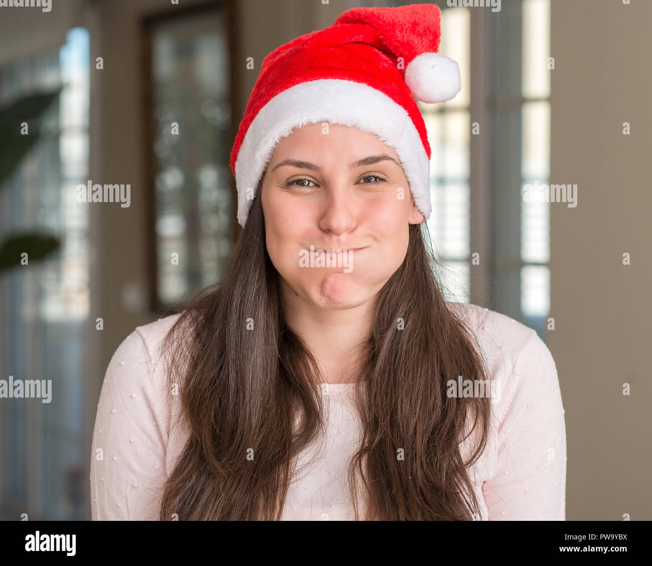 Beautiful Young Woman Wearing Santa Claus Hat At Home Puffing Cheeks