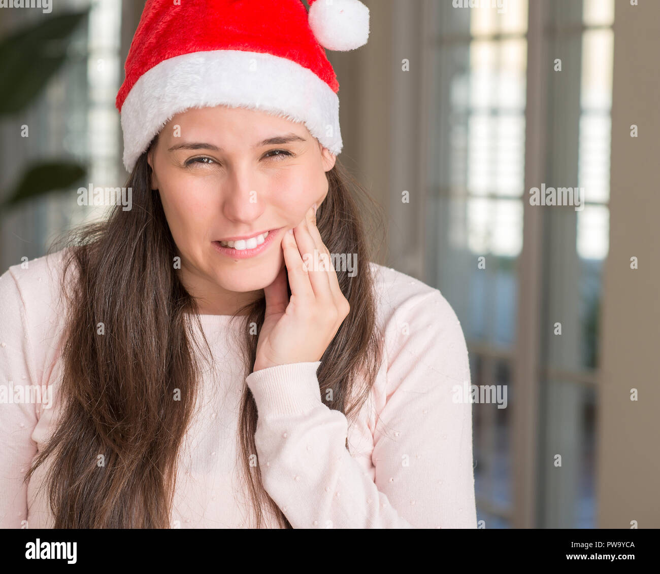 Beautiful young woman wearing Santa Claus hat at home touching mouth ...