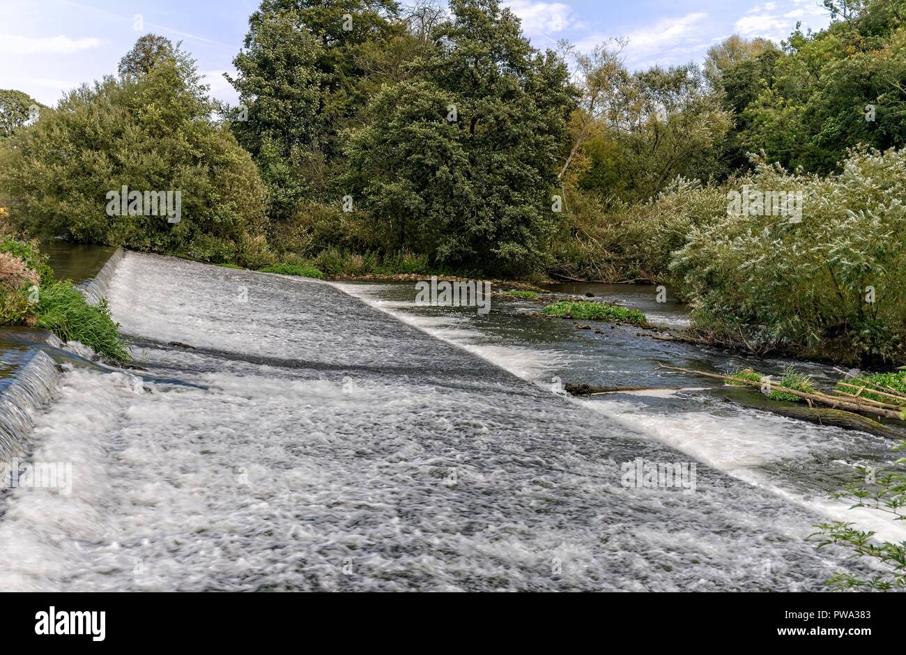 Water rushes down a weir to the river below. Trees of a woodland are on ...