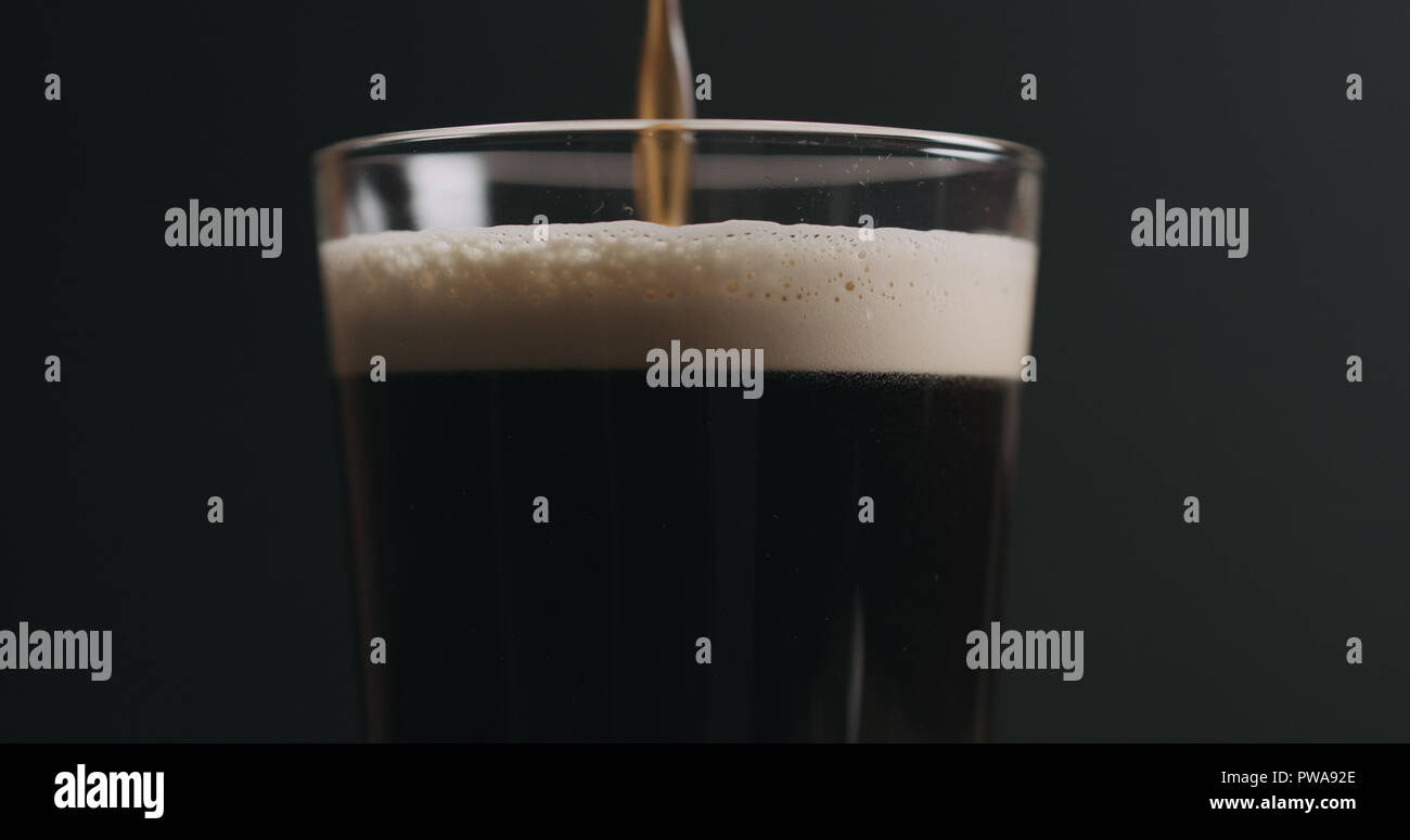 Closeup pour dark stout beer in pint glass over black background, wide ...