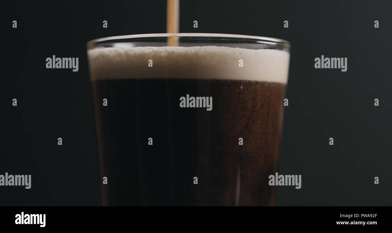 Closeup pour dark stout beer in pint glass over black background, wide ...