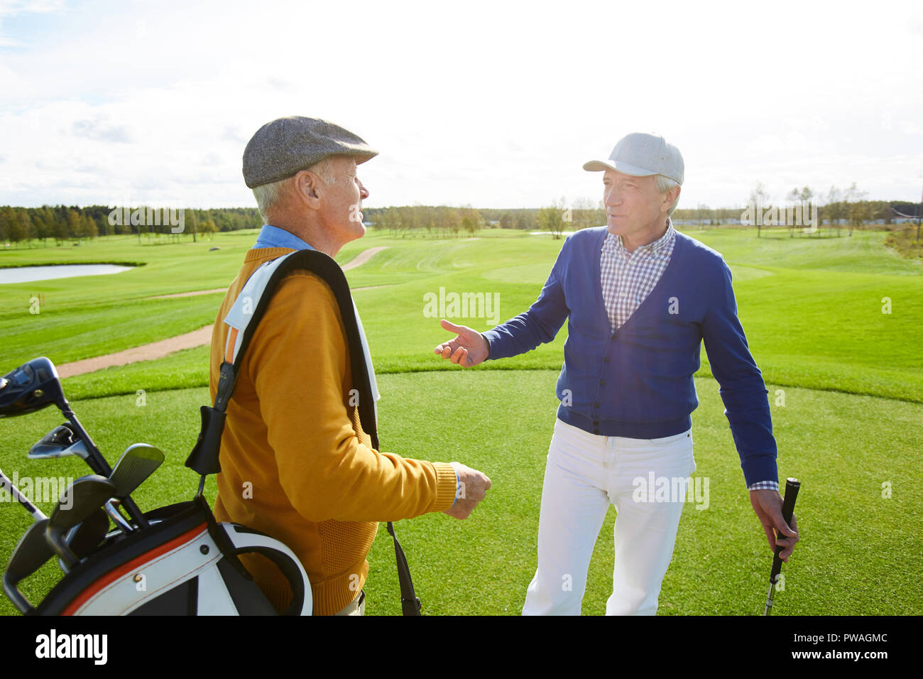 Two senior men with golf clubs talking in the middle of green field ...
