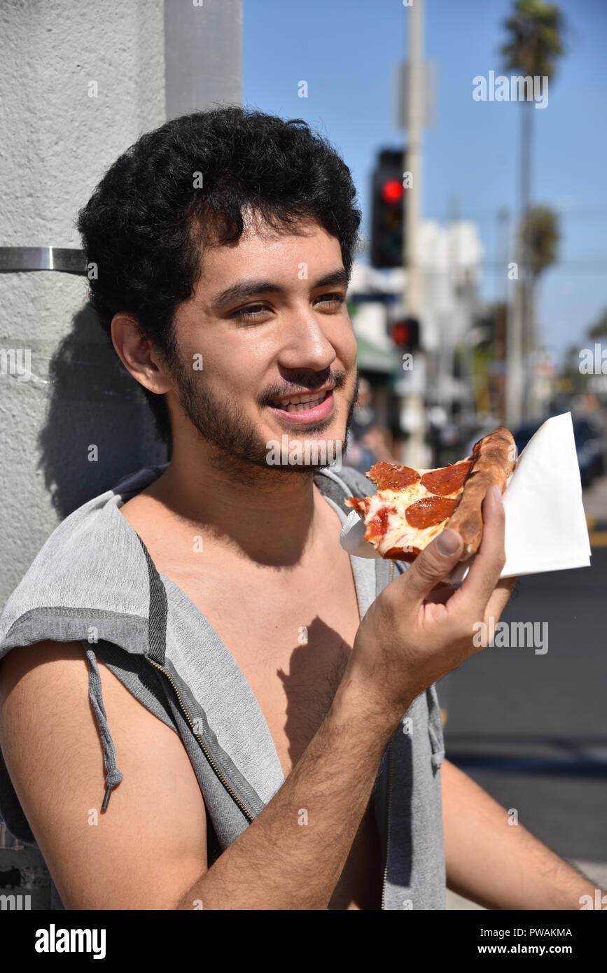 A happy young man leaning against a lamp post eating a slice of ...