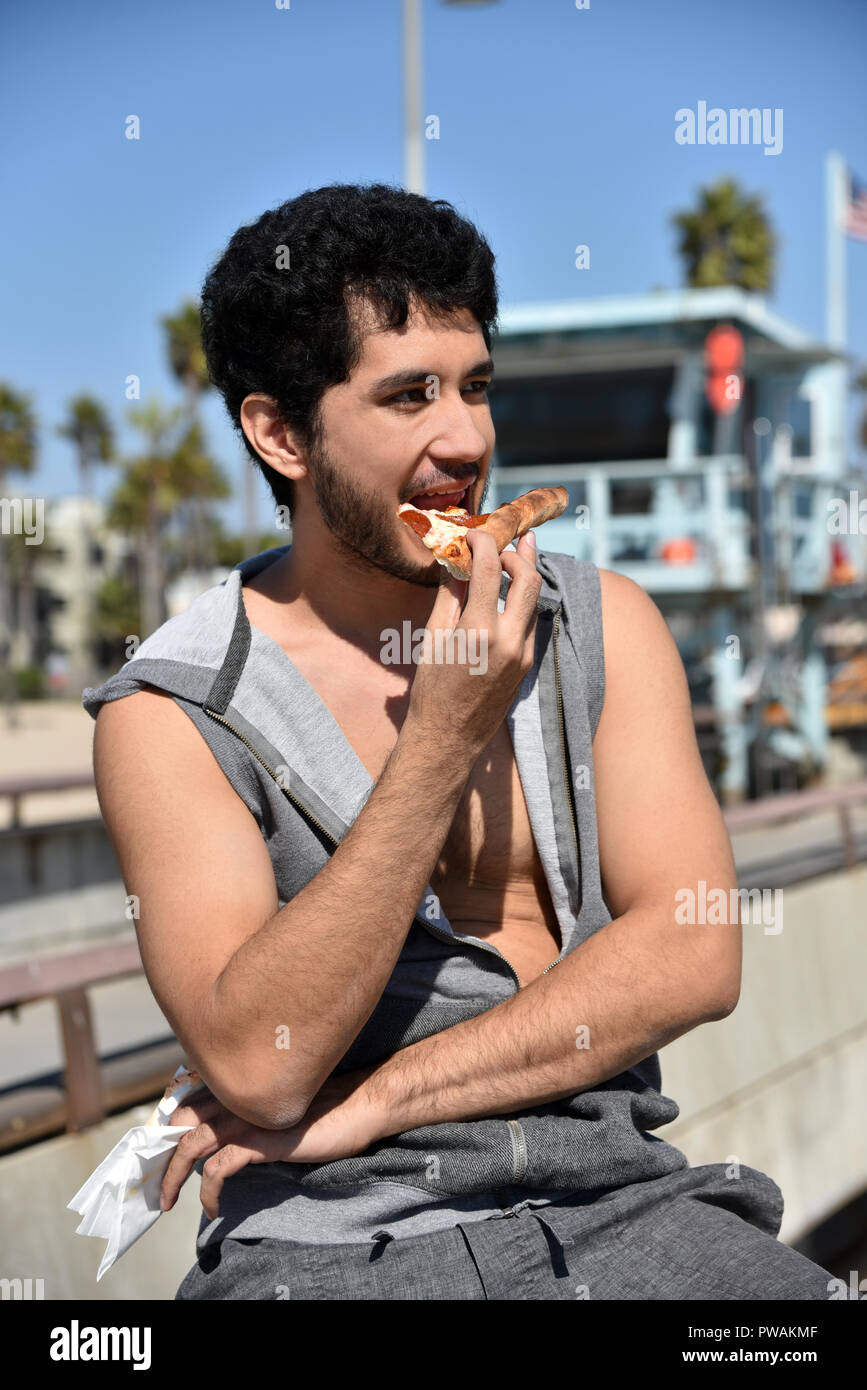 A happy multi-ethnic young man eating a slice of pizza on the pier by a ...