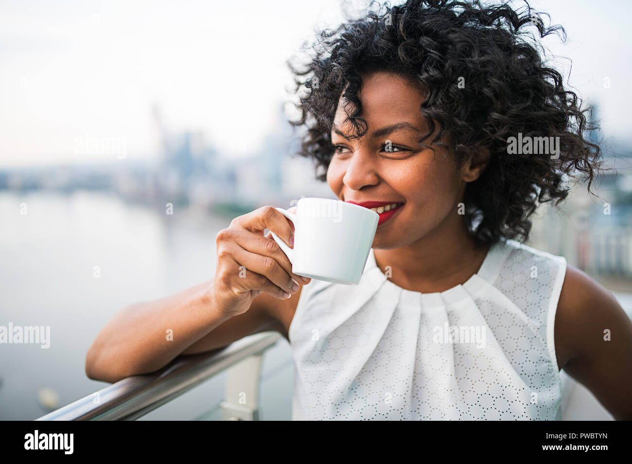 A portrait of a black woman standing on a terrace, drinking coffee ...