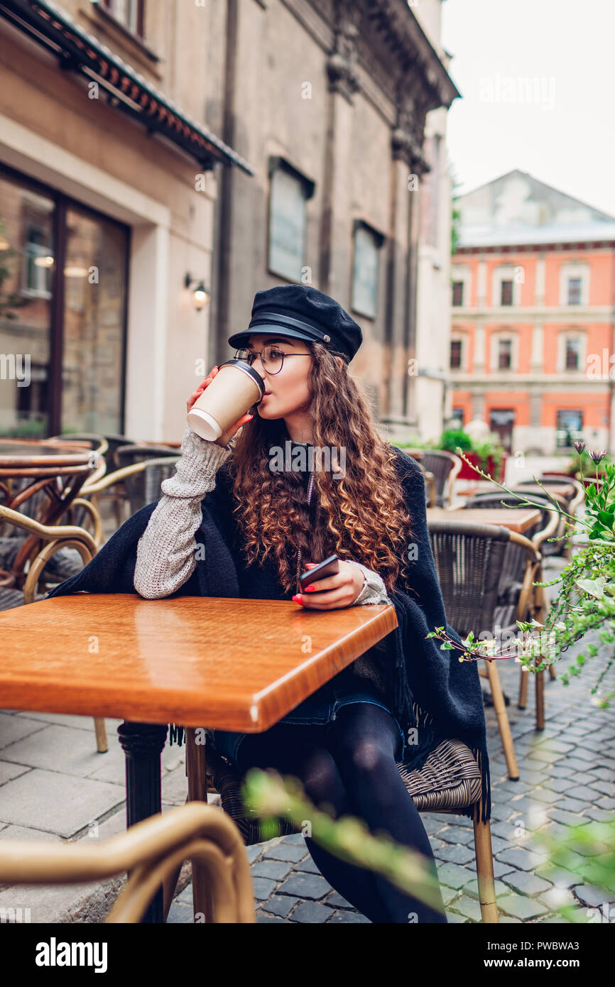 Stylish young woman having coffee in outdoor cafe while using ...