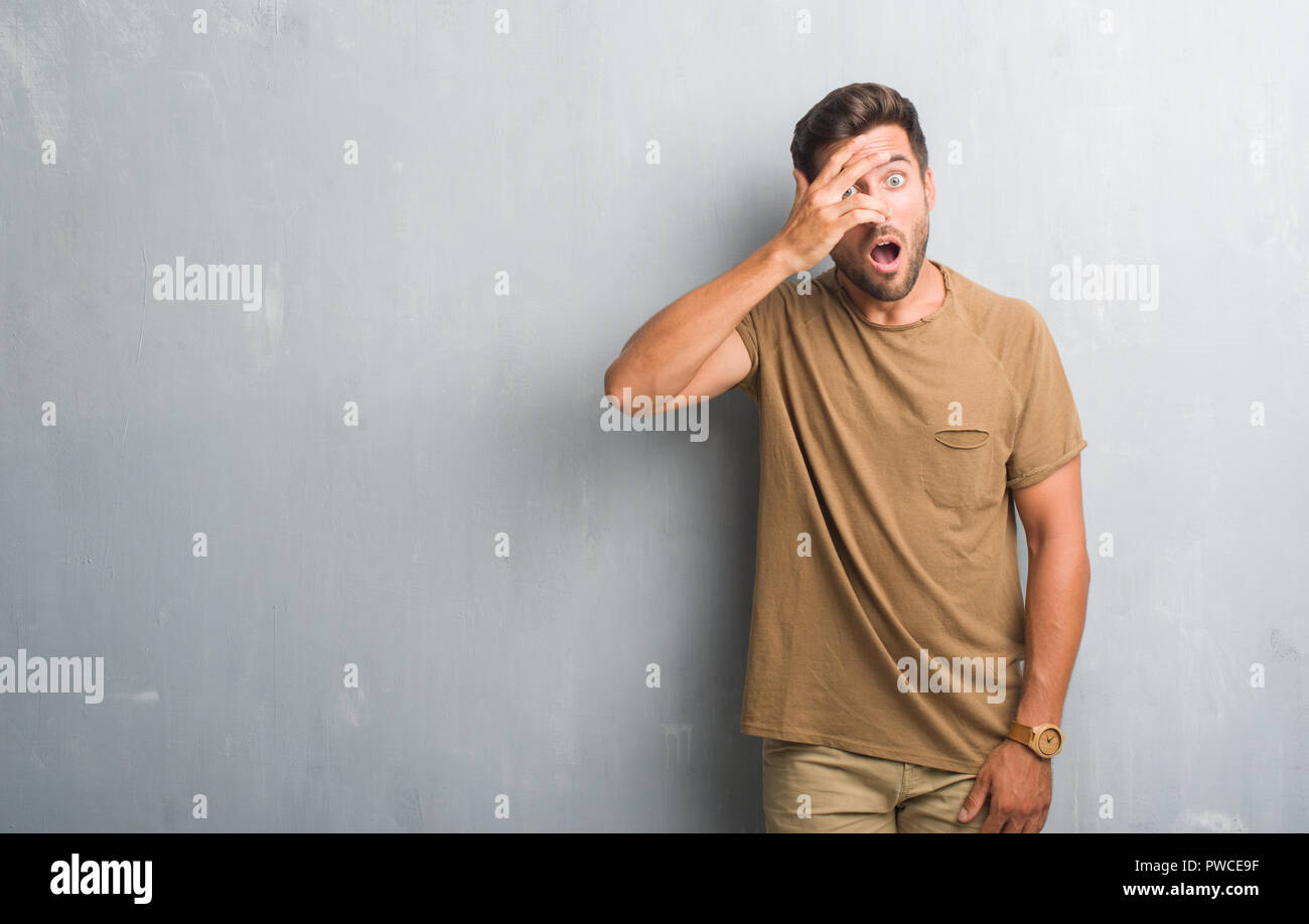 Handsome young man over grey grunge wall peeking in shock covering face ...
