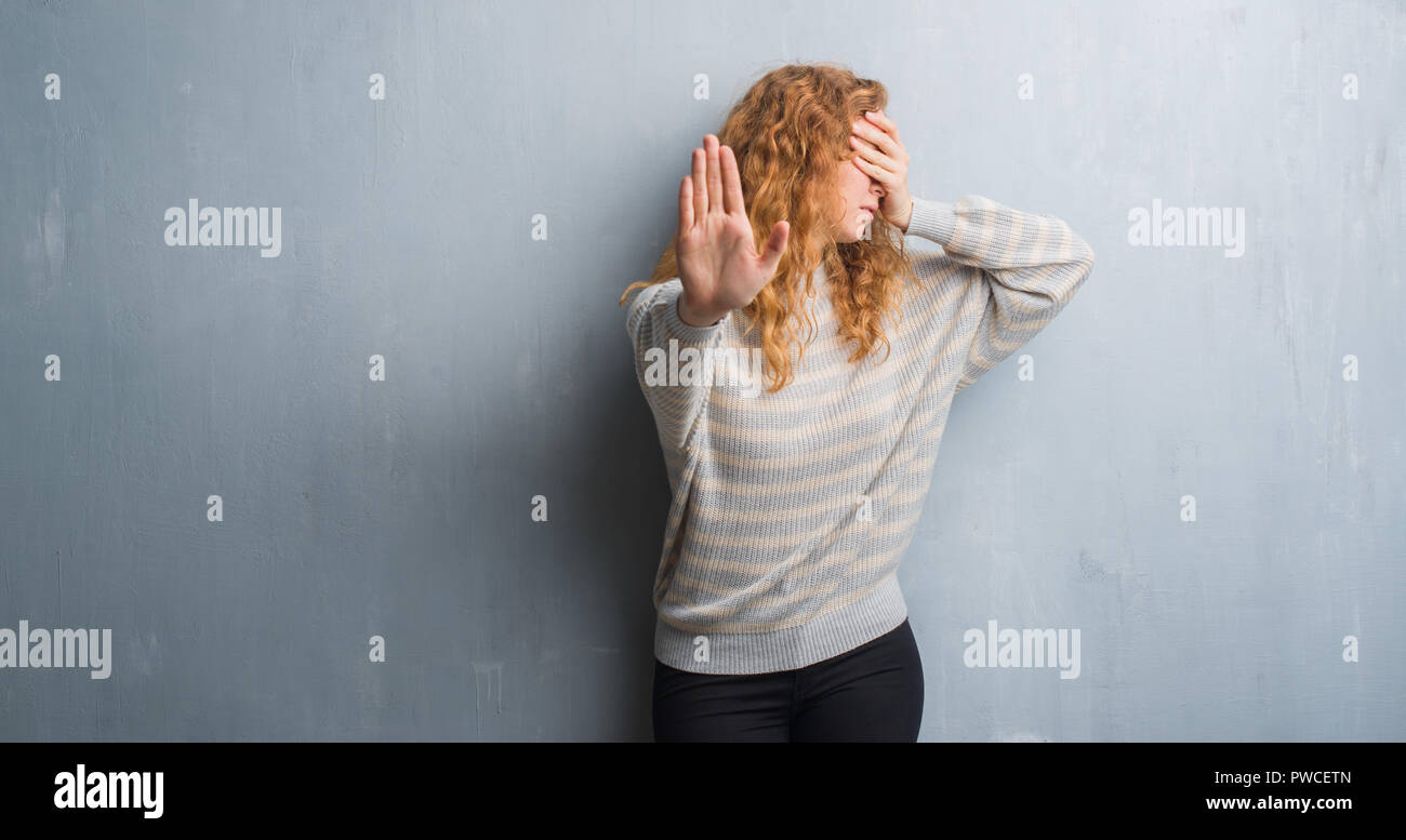 Young redhead woman over grey grunge wall covering eyes with hands and ...