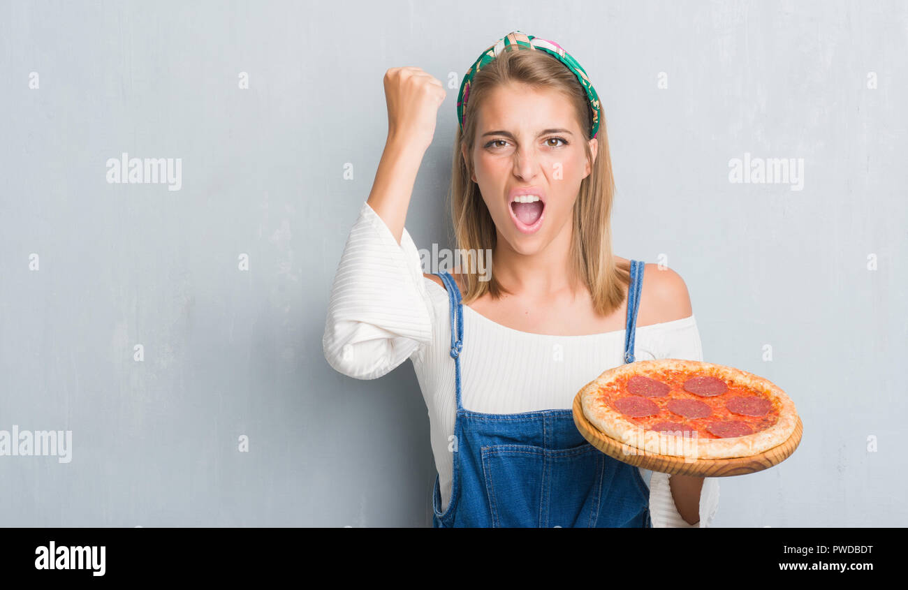 Beautiful young woman over grunge grey wall eating pepperoni pizza ...