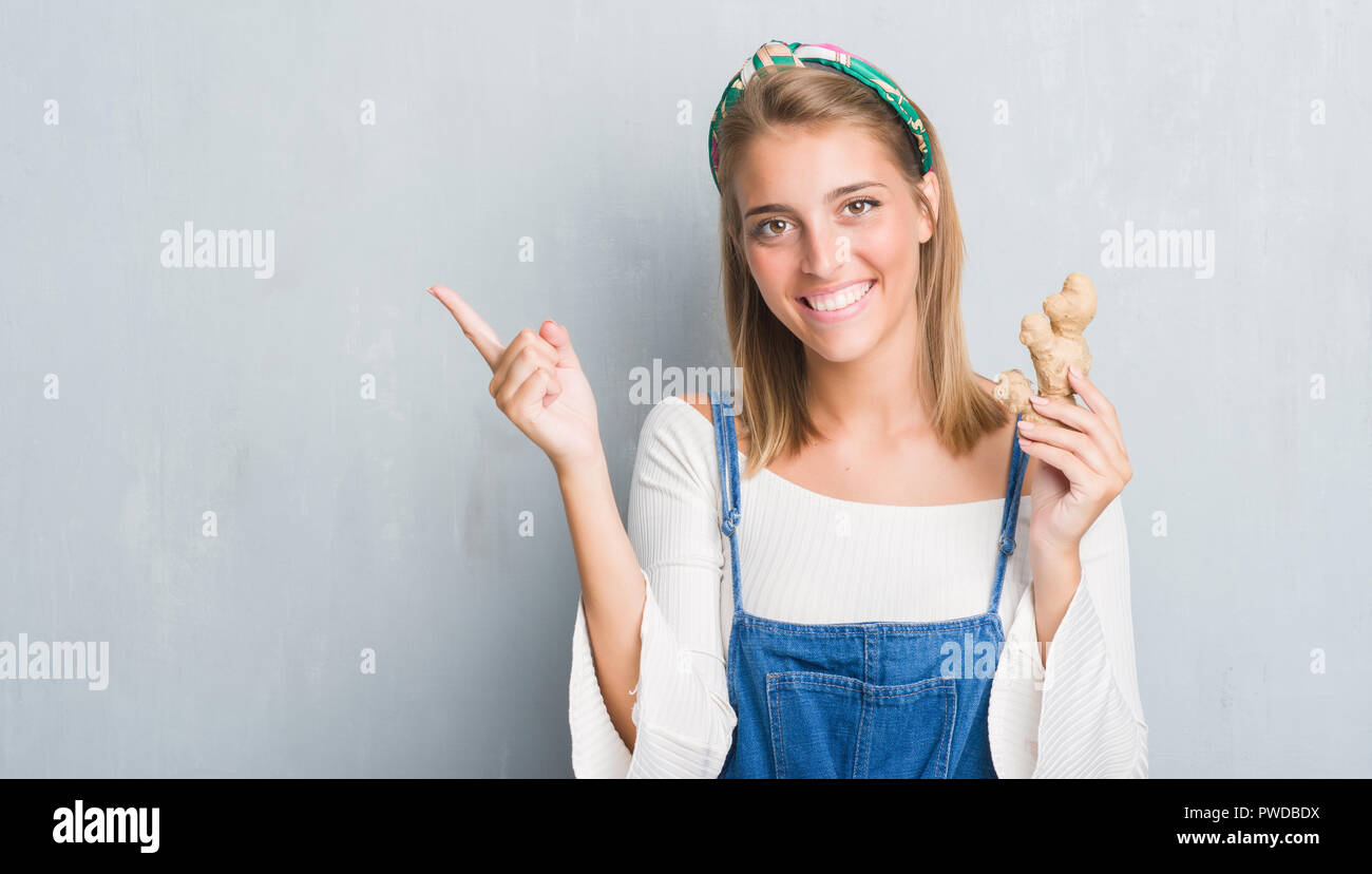 Beautiful young woman over grunge grey wall holding fresh ginger very ...
