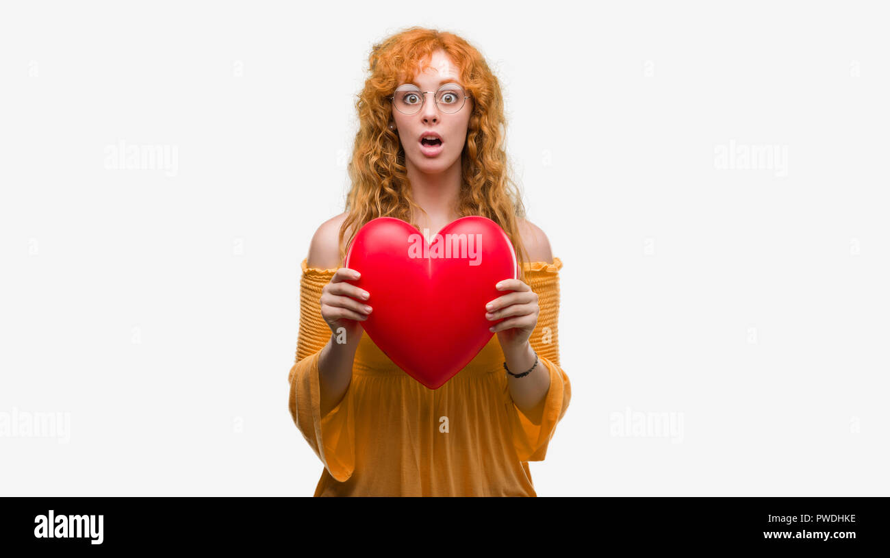 Young redhead woman in love holding red heart scared in shock with a ...