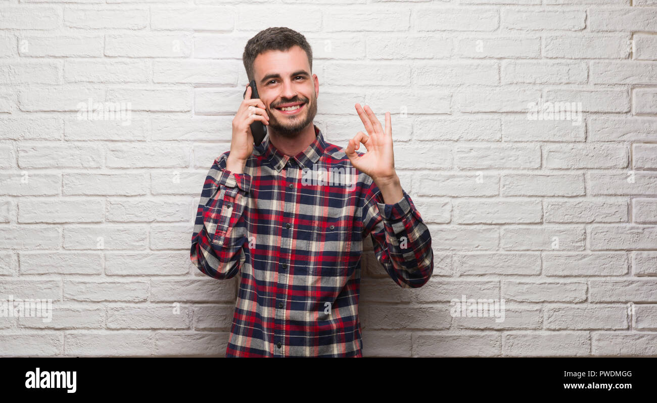 Young adult man talking on the phone standing over white brick wall ...