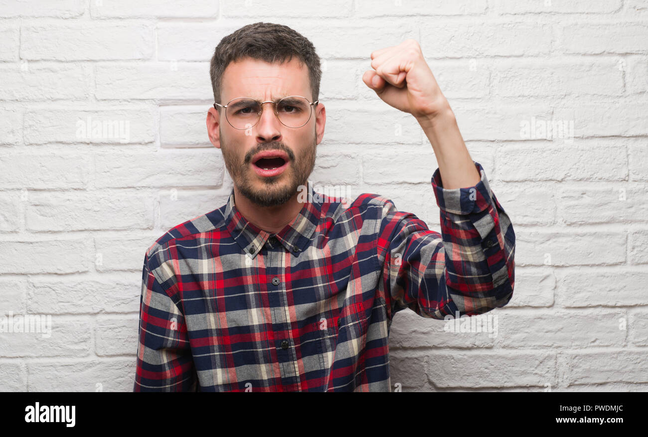 Young adult man standing over white brick wall annoyed and frustrated ...