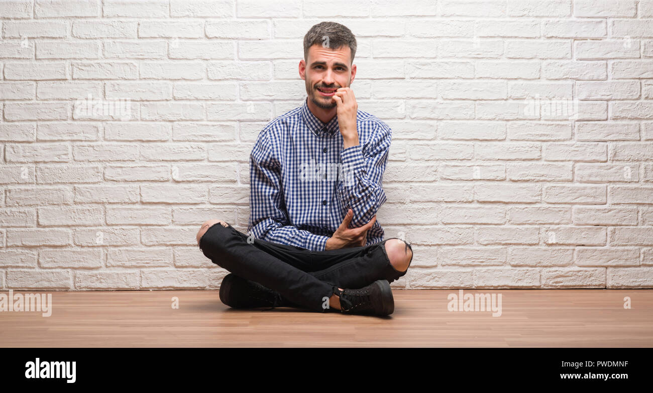 Young adult man sitting over white brick wall looking stressed and ...