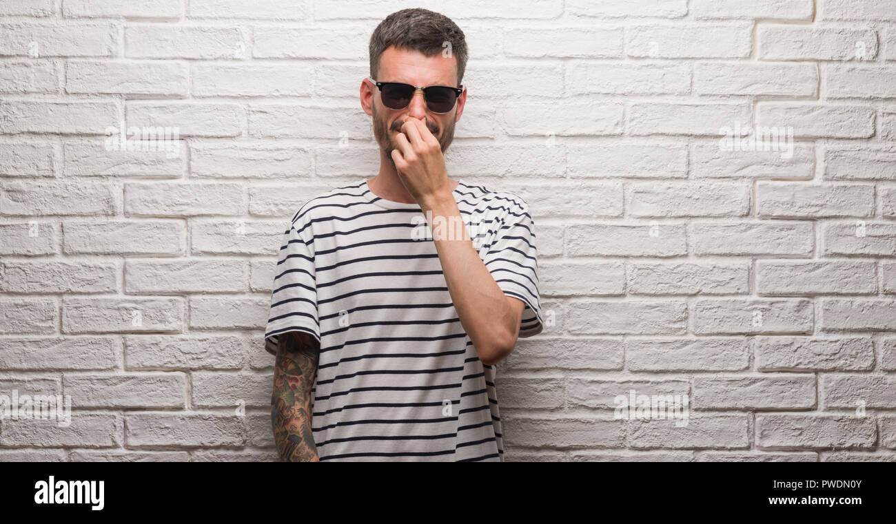 Young adult man wearing sunglasses standing over white brick wall ...