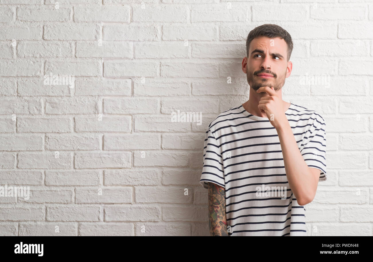 Young adult man standing over white brick wall serious face thinking ...
