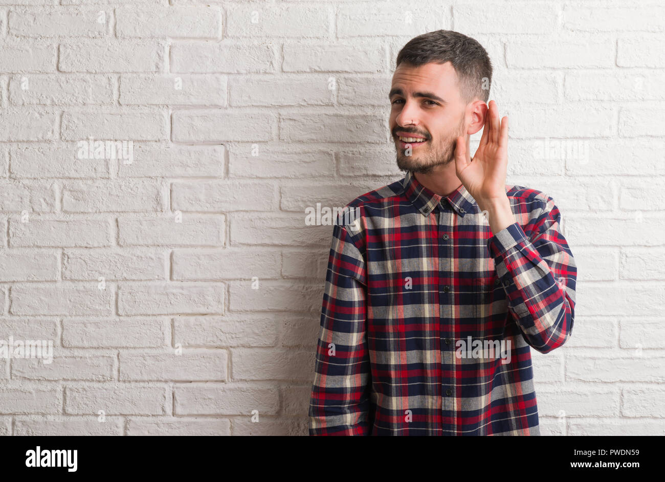 Young adult man standing over white brick wall smiling with hand over ...