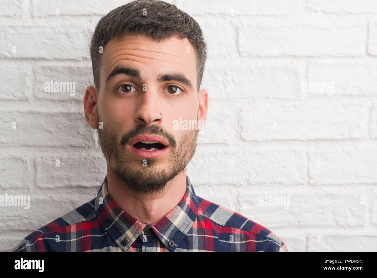 Young adult man standing over white brick wall scared in shock with a ...