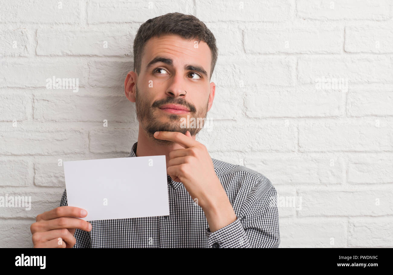 Young adult man over brick wall holding blank paper serious face ...