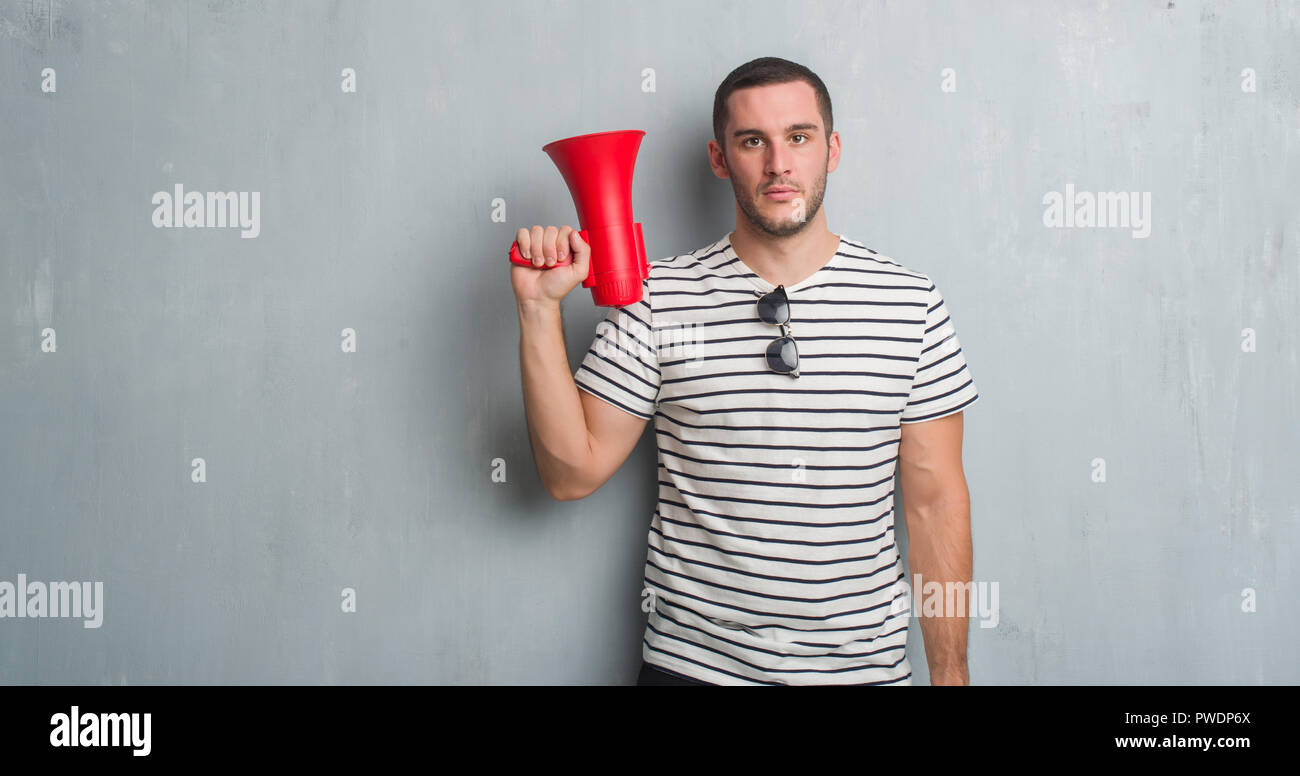 Young caucasian man over grey grunge wall shouting trough megaphone ...