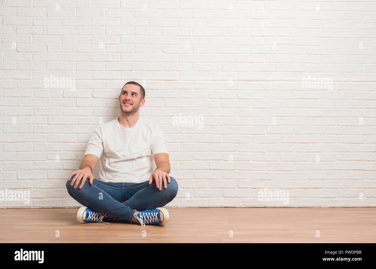 Young caucasian man sitting on the floor over white brick wall smiling ...
