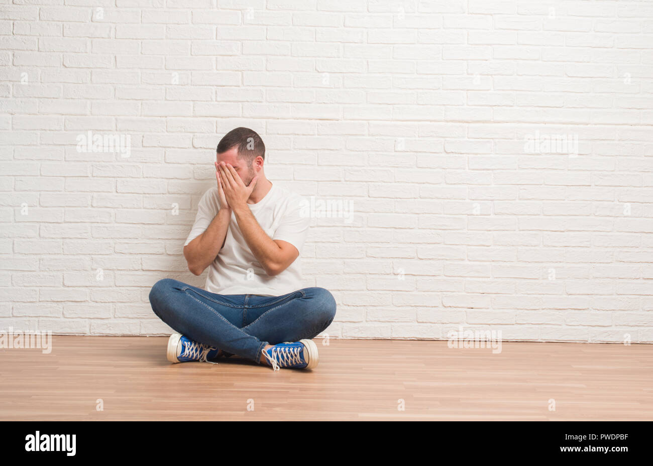 Young caucasian man sitting on the floor over white brick wall with sad ...