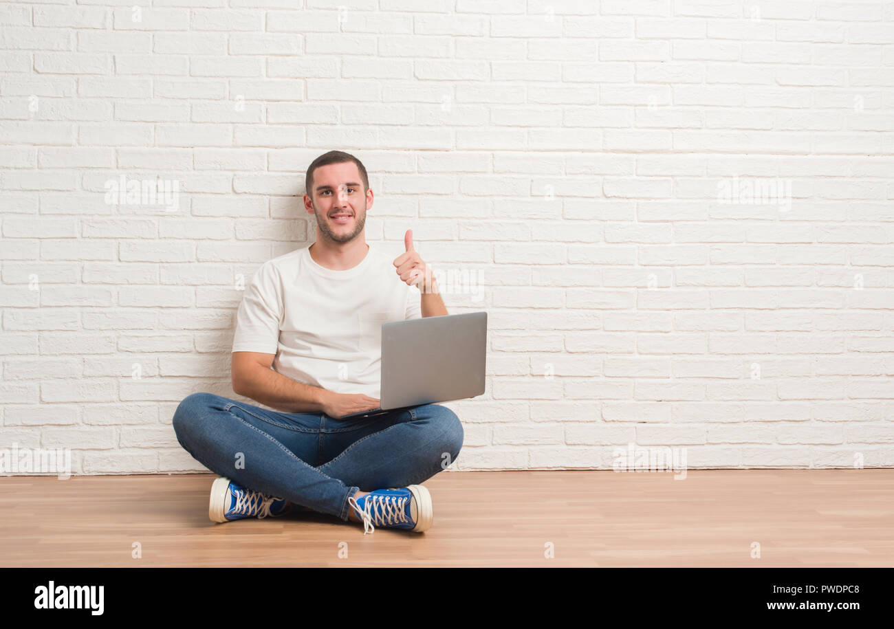 Young caucasian man sitting over white brick wall using computer laptop ...