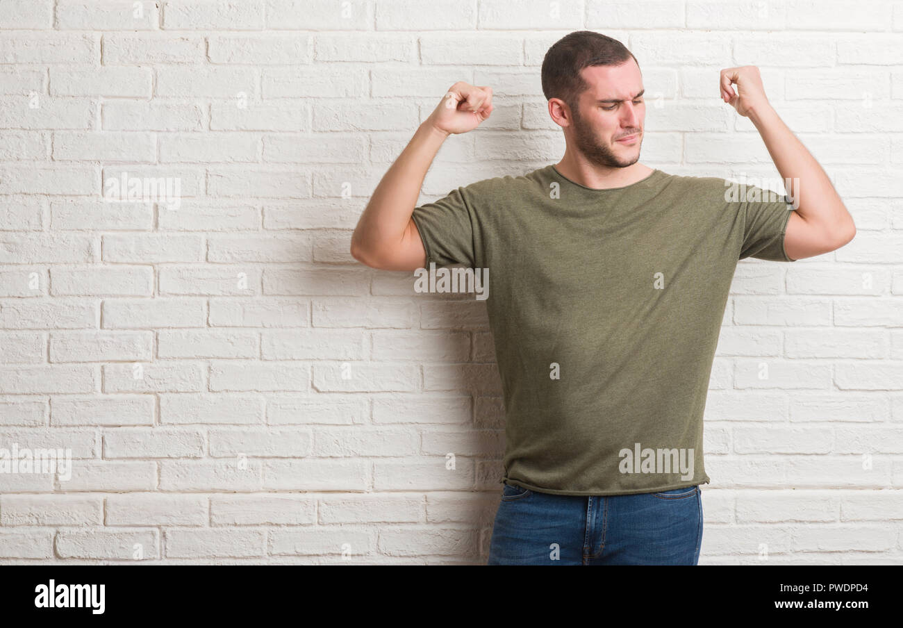 Young caucasian man standing over white brick wall showing arms muscles ...