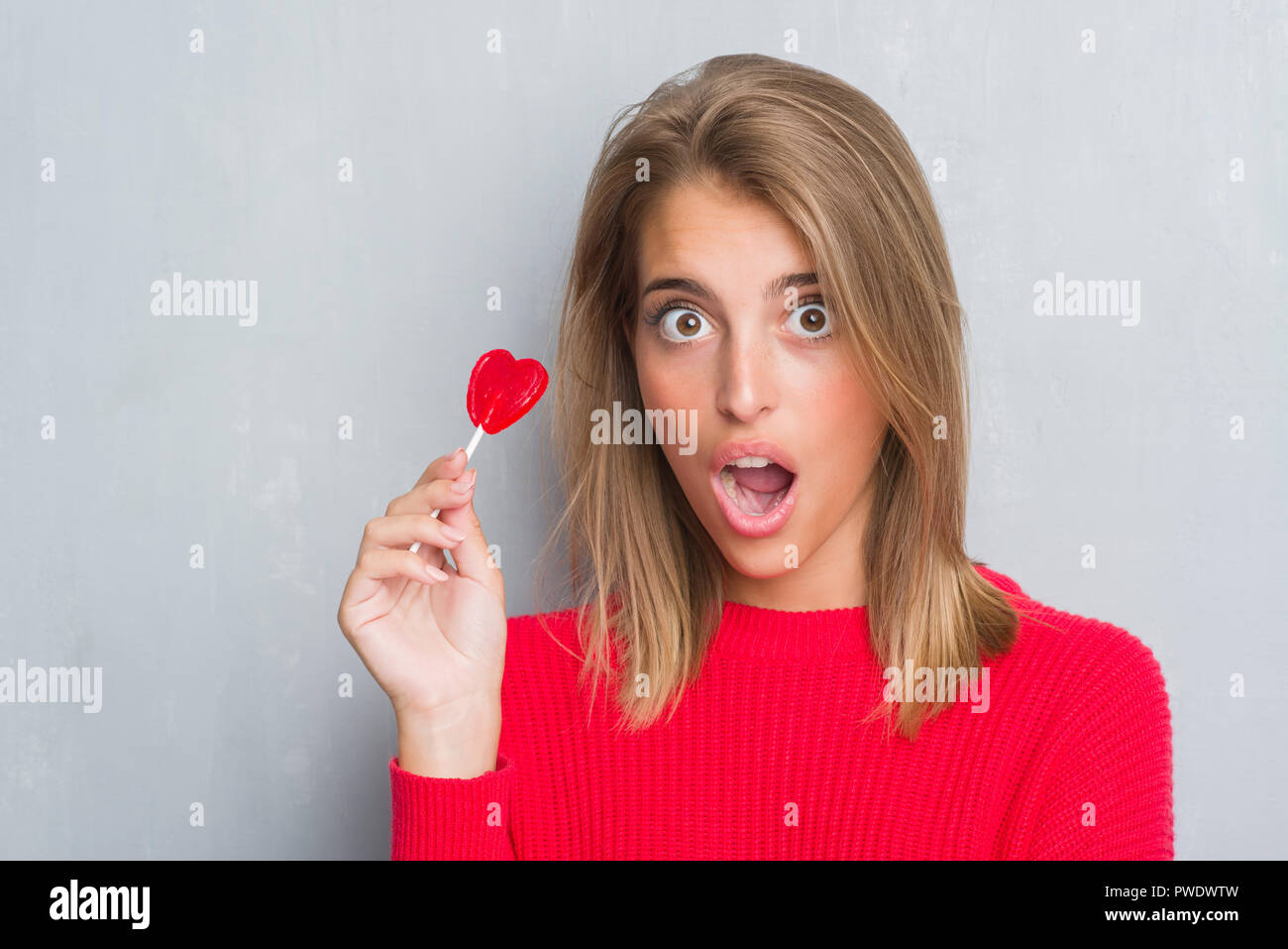 Beautiful young woman over grunge grey wall eating red heart lollipop ...