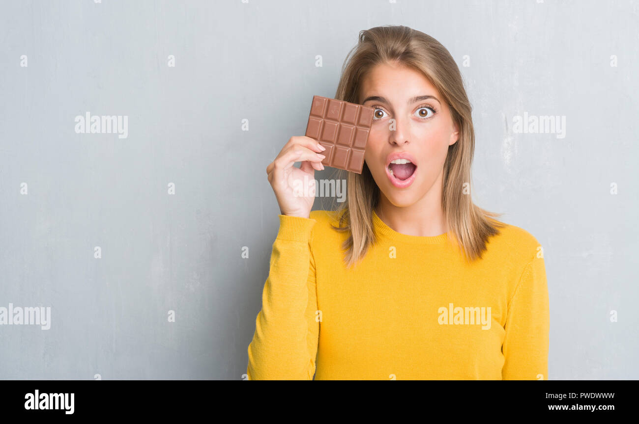 Beautiful young woman over grunge grey wall eating chocolate bar scared ...