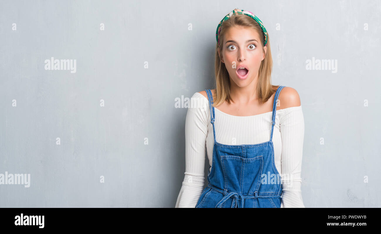 Beautiful young woman standing over grunge grey wall scared in shock ...