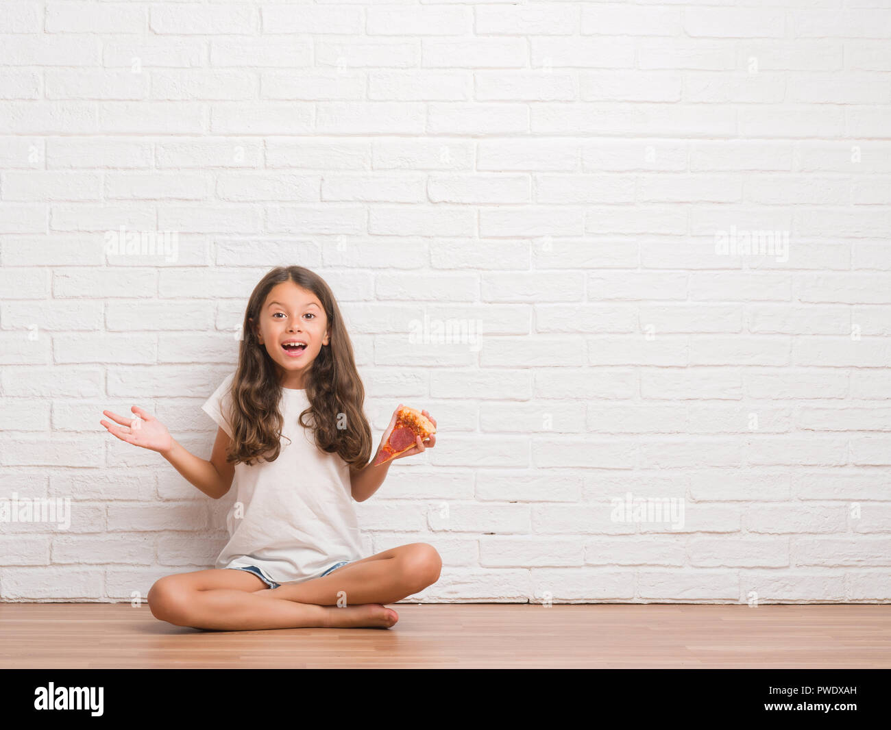 Young hispanic kid sitting on the floor over white brick wall eating ...