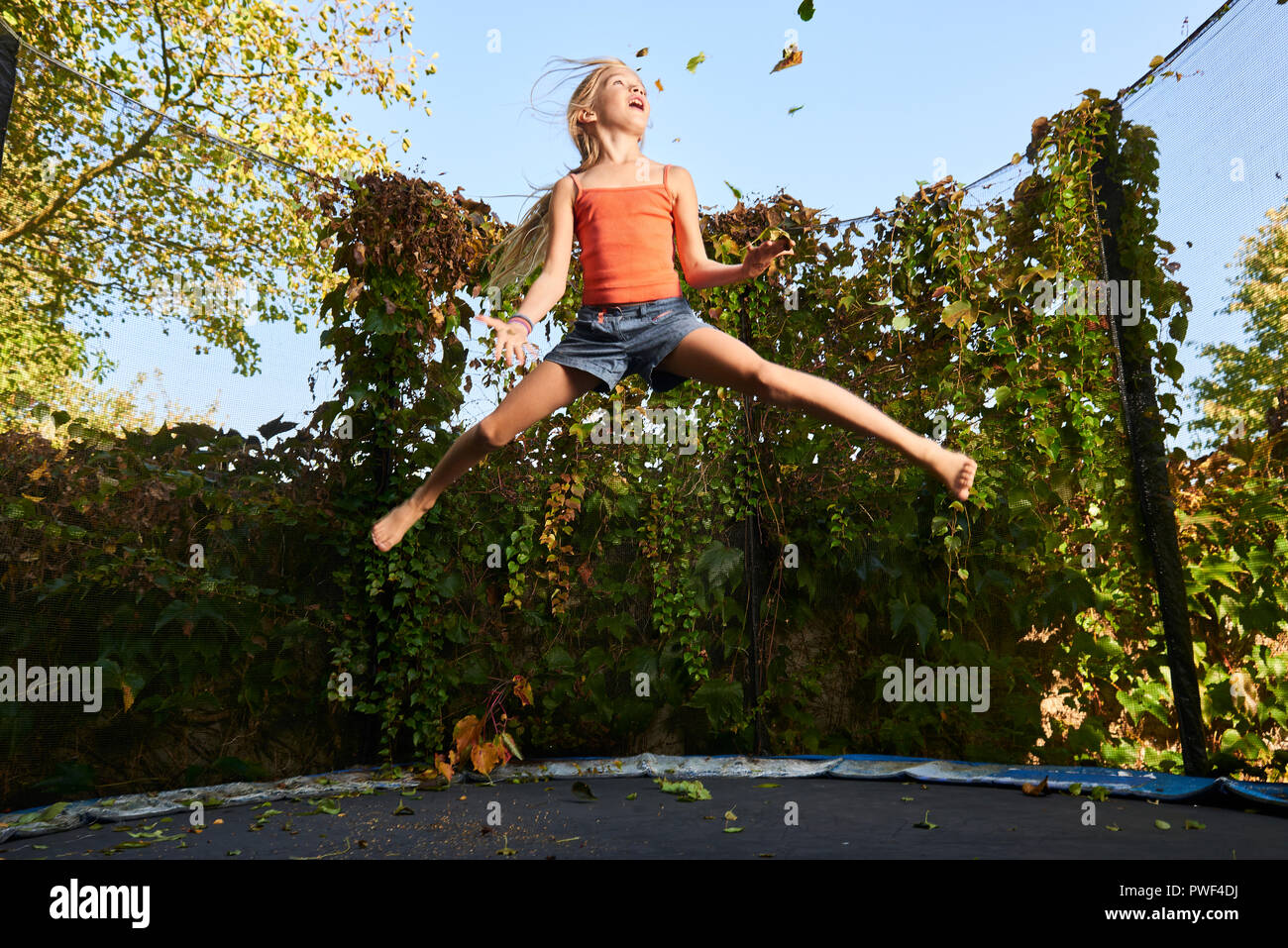 Child cute blond girl playing and jumping on trampoline with greenery ...