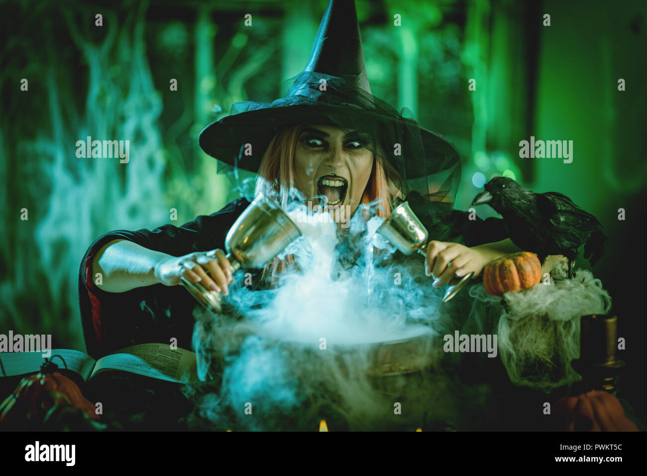 A witch in foggy surrounding holds two goblets above boiling water for ...