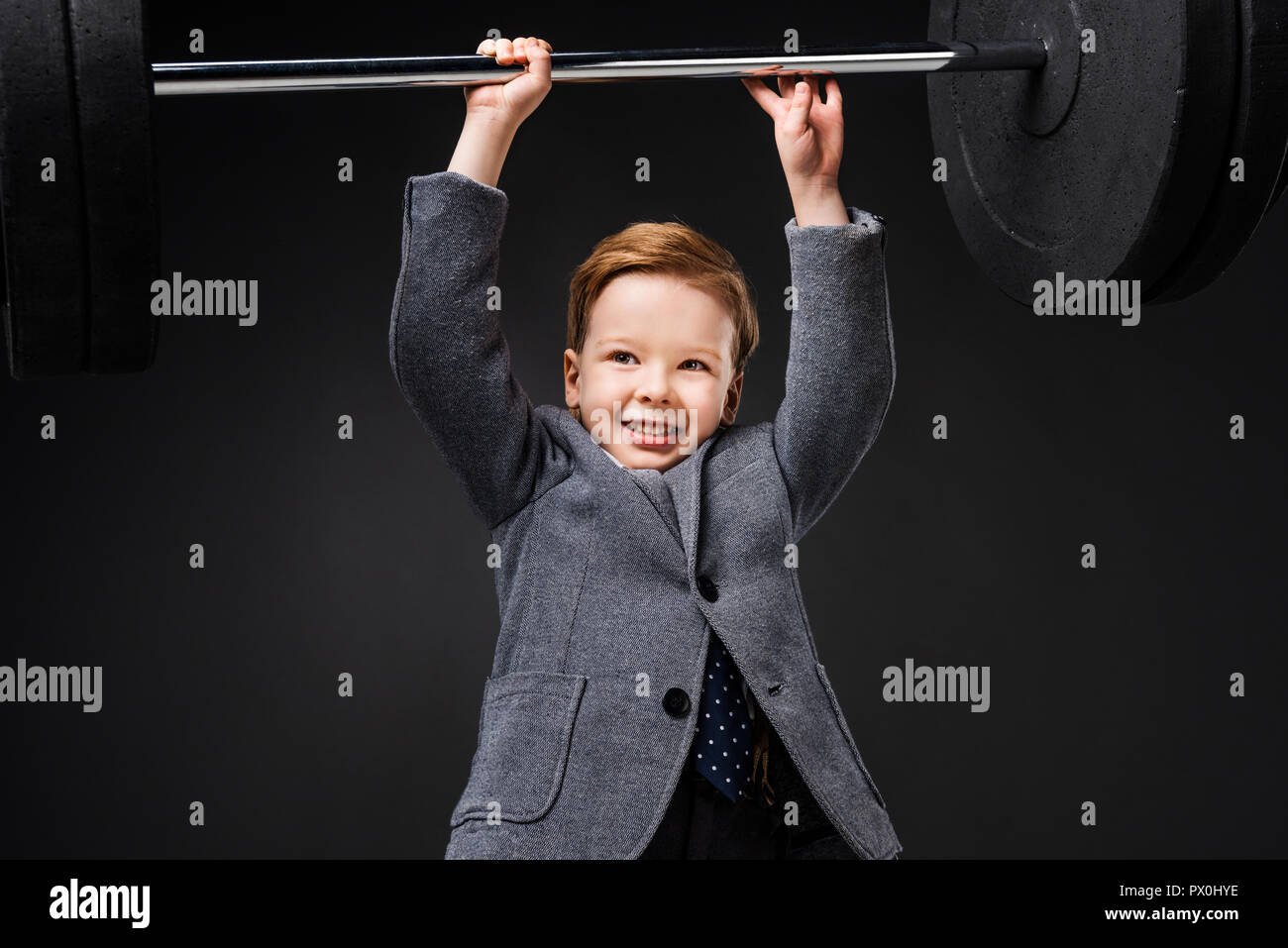 strong smiling little boy in suit lifting barbell isolated on grey ...