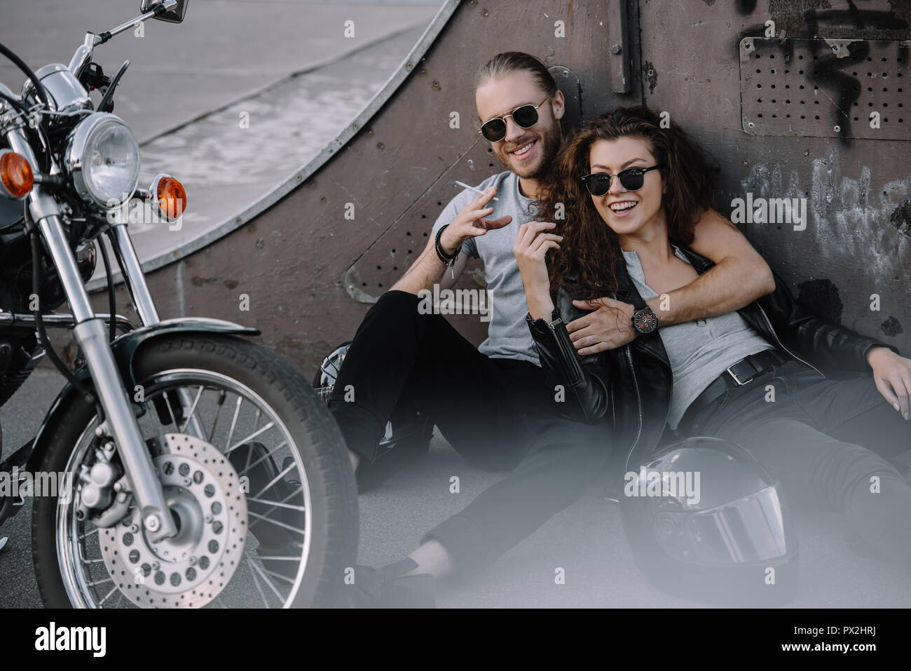smiling couple of bikers embracing and smoking on asphalt with chopper ...
