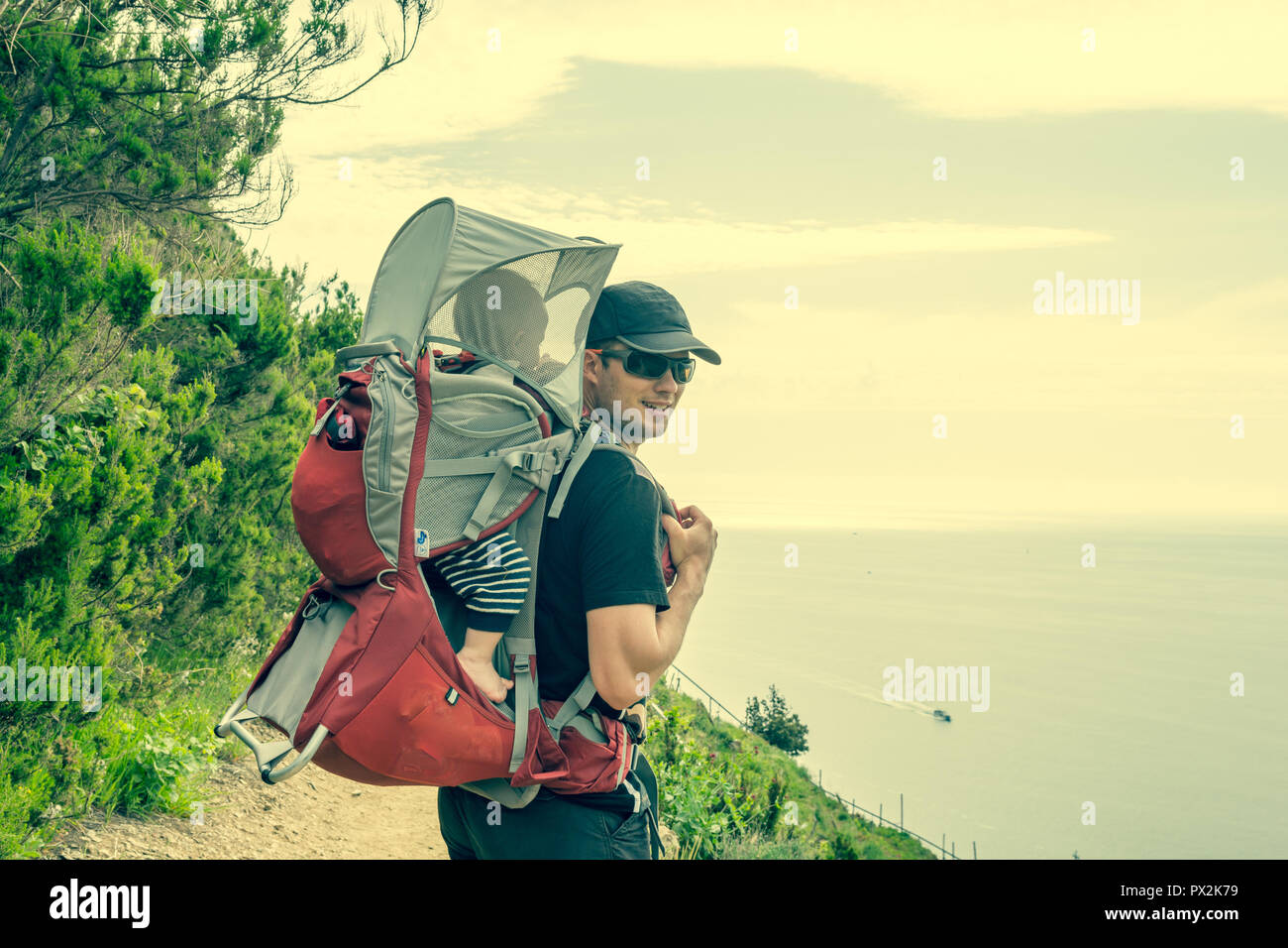 Active father carrying a baby carrier and walking along a coastal path ...