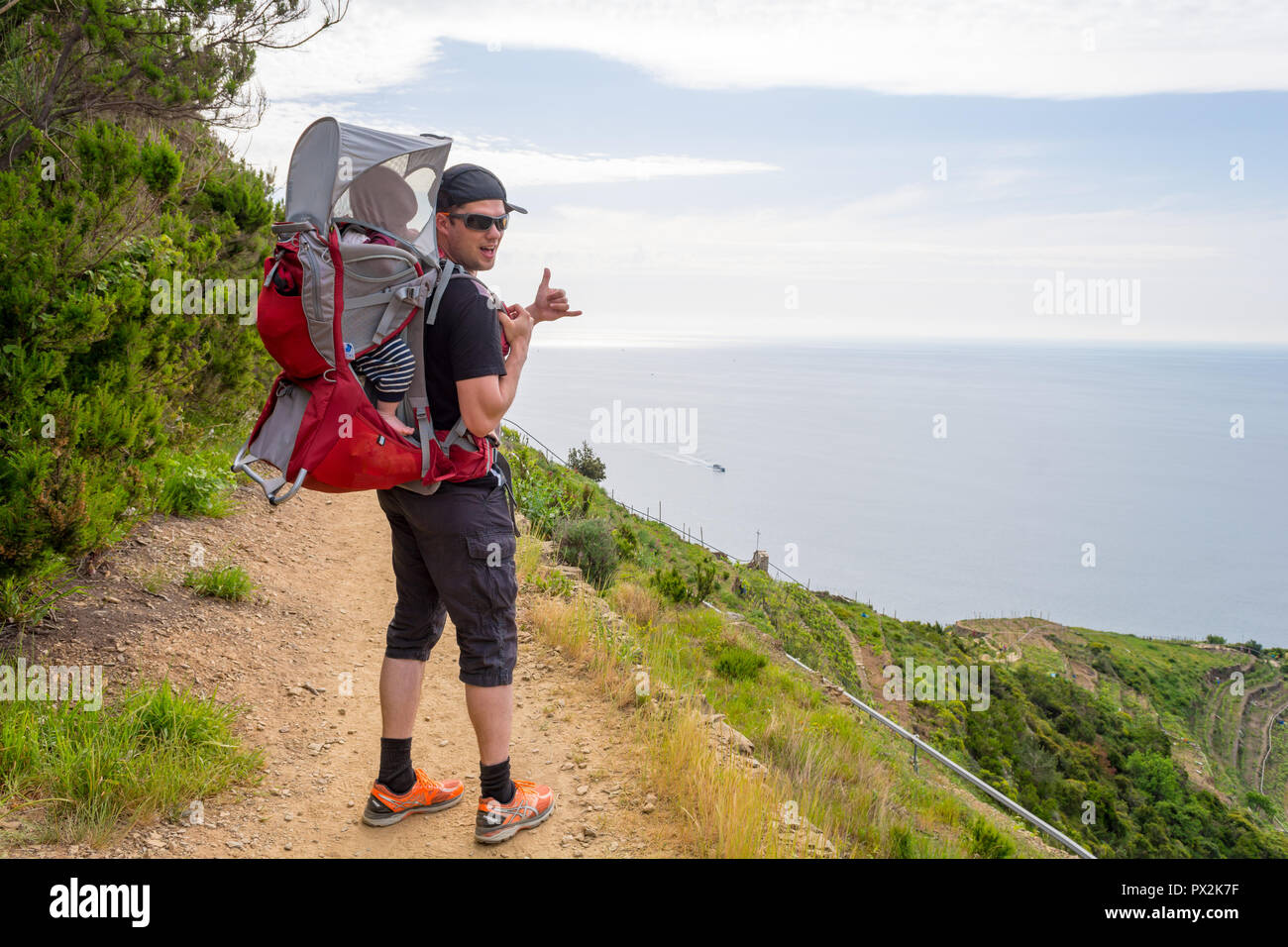 Active father carrying a baby carrier and walking along a coastal path ...