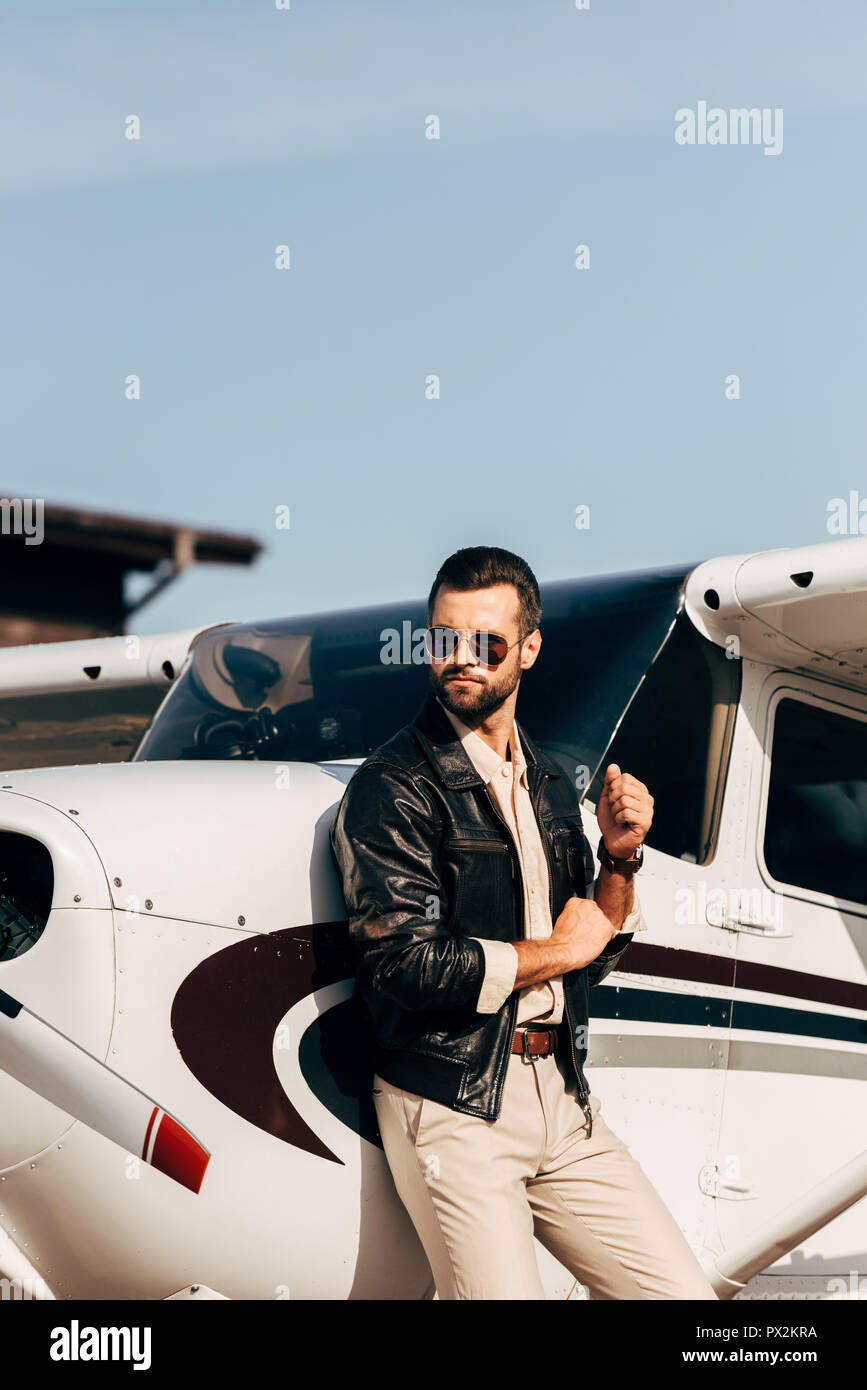 male pilot in leather jacket and sunglasses looking away near aircraft ...