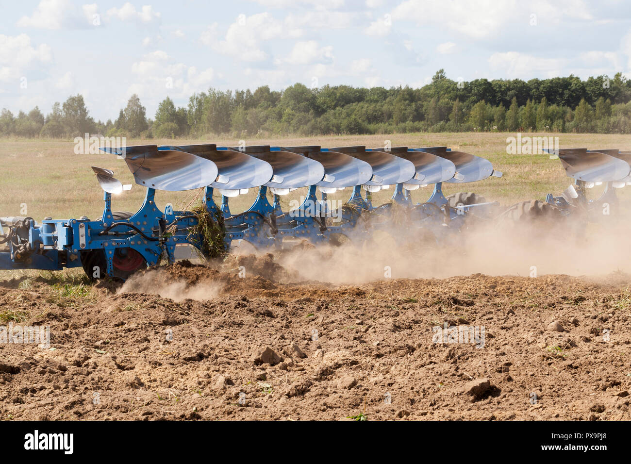 large blue plow plows the soil on the field, a lot of dust from the dry ...