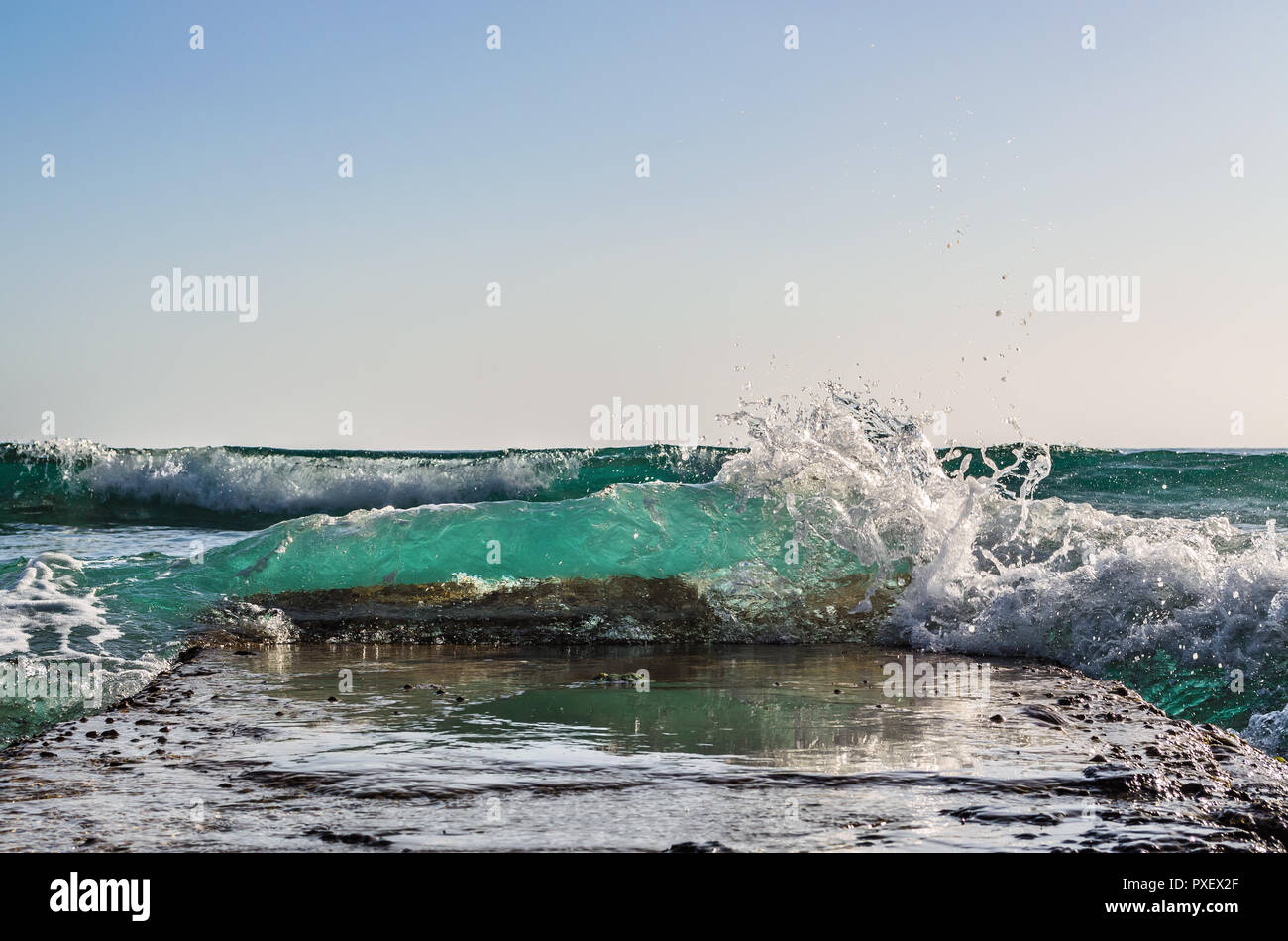 A turquoise wave is breaking on a rocky pier with clean sky Stock Photo ...