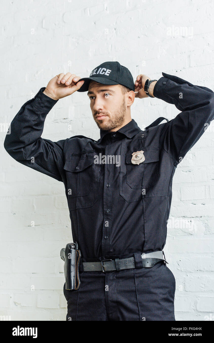 handsome young policeman putting on cap in front of white brick wall ...