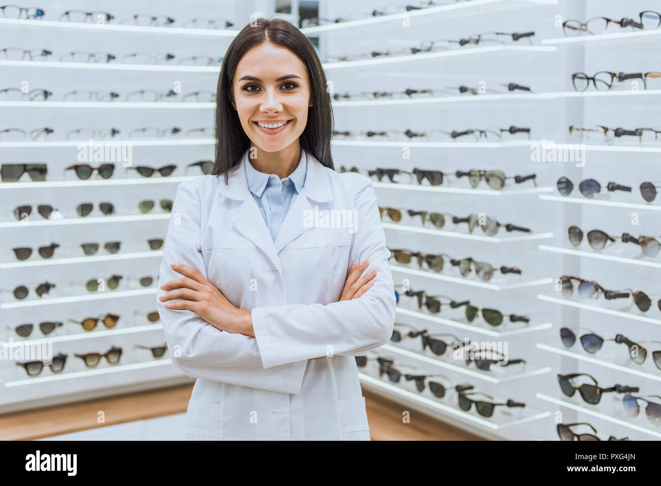 professional smiling optometrist posing with crossed arms near shelves ...