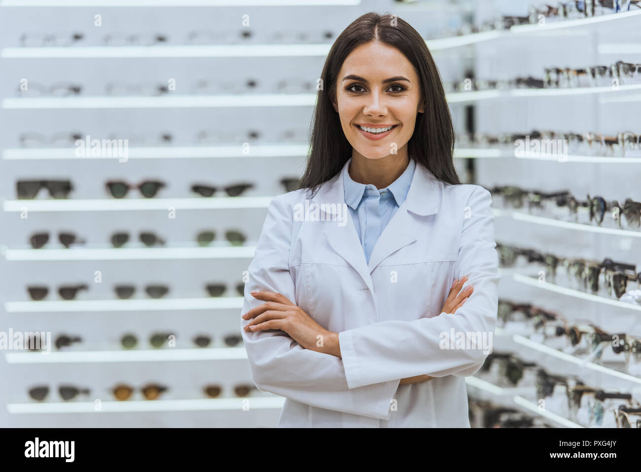 professional confident optician posing with crossed arms near shelves ...